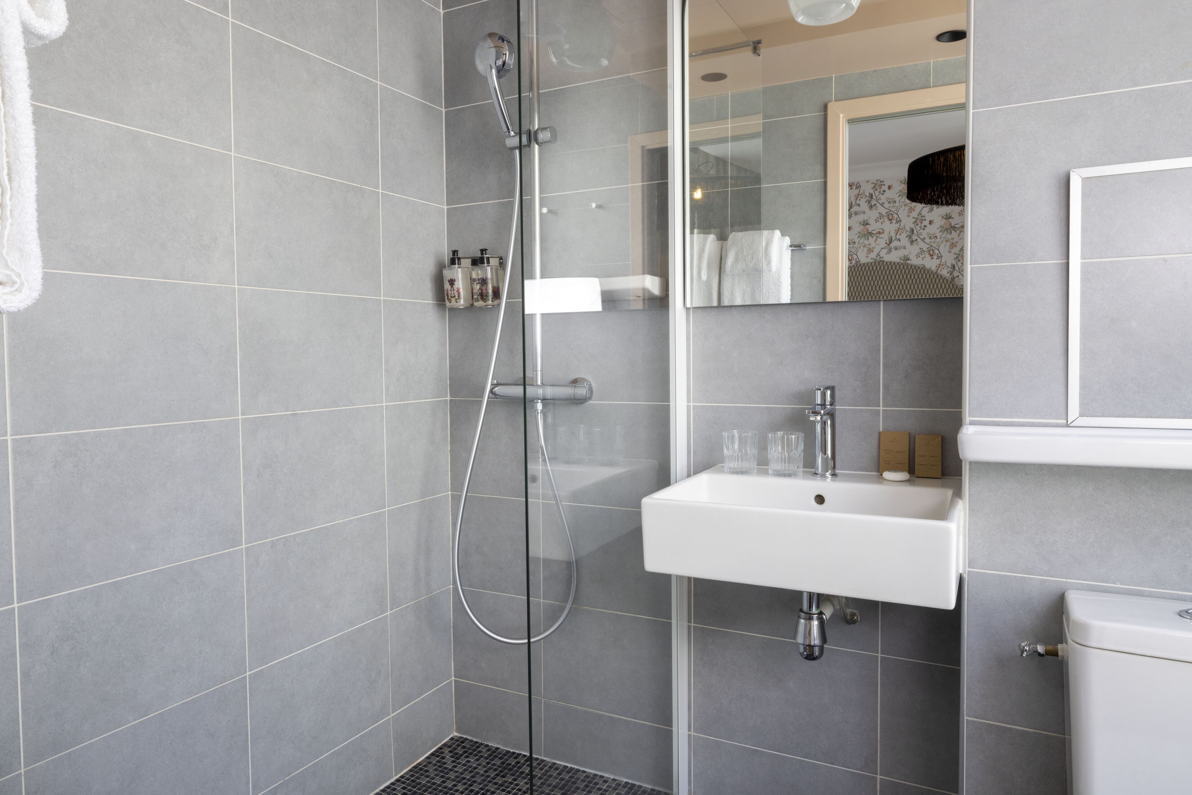 Casa de banho moderna com azulejos cinzentos, cabine de duche em vidro, lavatório branco e sanita.