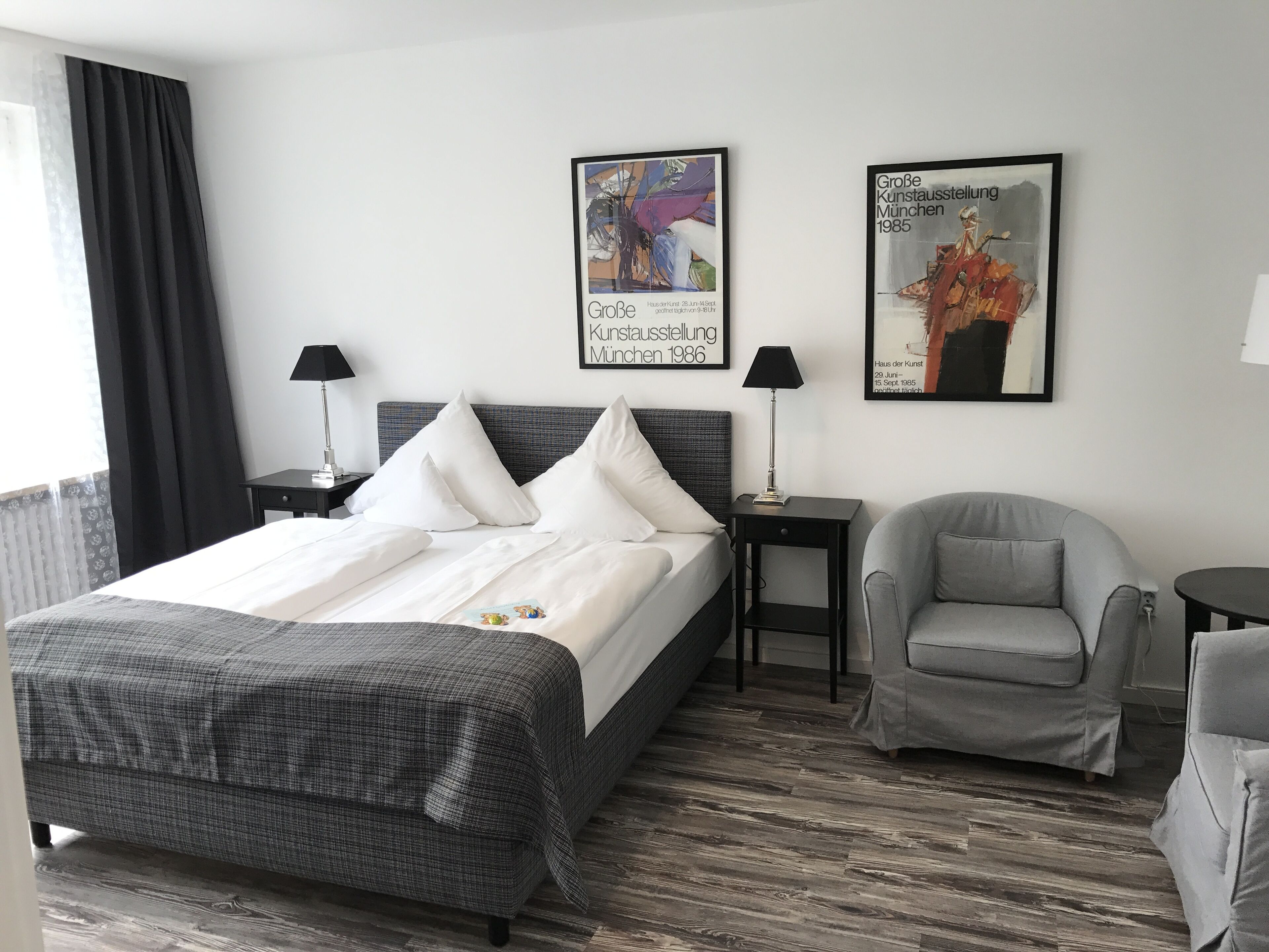 Quarto de hotel com uma cama grande, uma poltrona cinza, duas mesas de cabeceira e dois quadros emoldurados na parede.