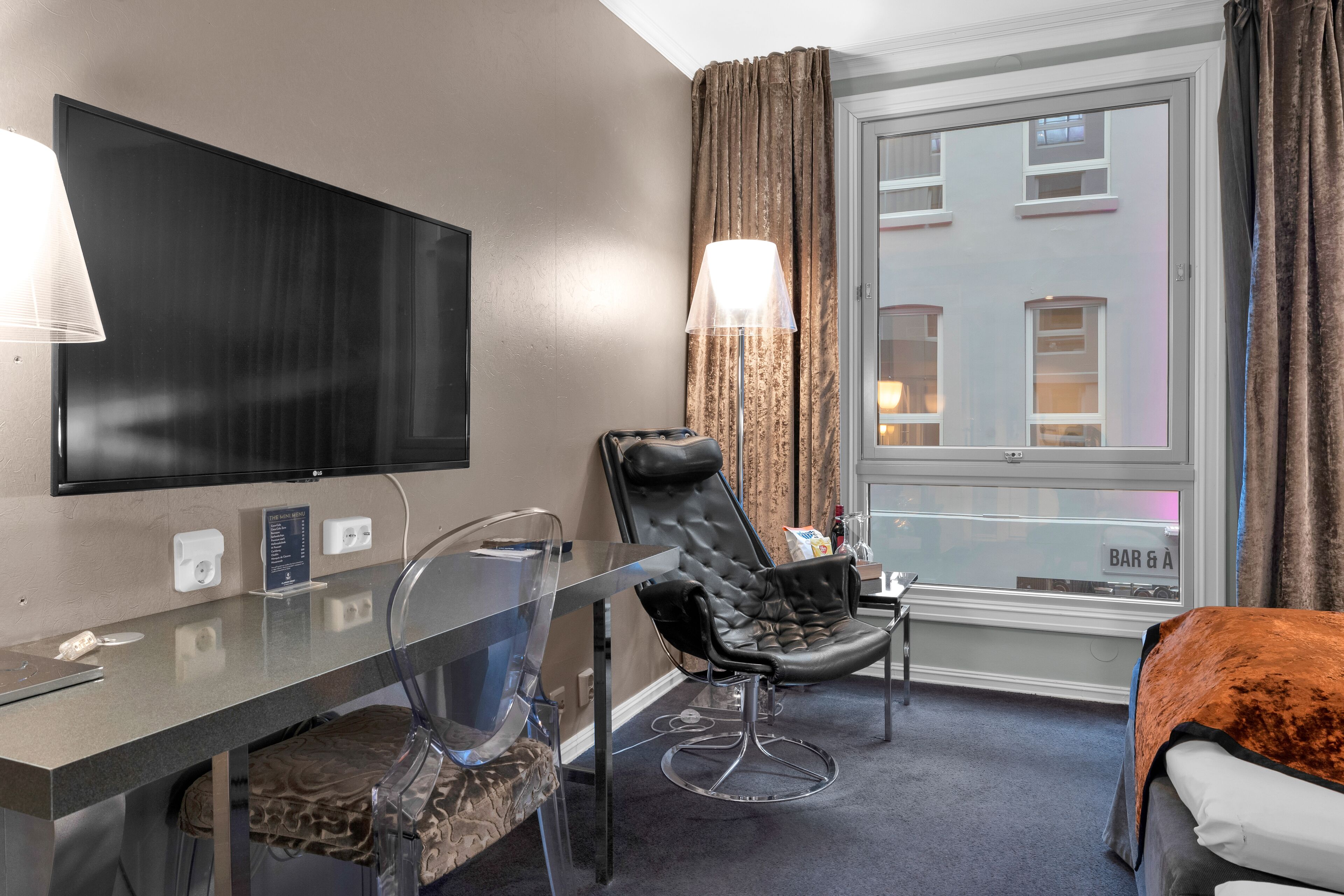 Quarto de hotel com televisão, escrivaninha com cadeira, cama e janela com cortinas.