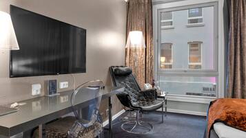 Quarto de hotel com televisão, escrivaninha com cadeira, cama e janela com cortinas.