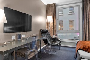 Quarto de hotel com televisão, escrivaninha com cadeira, cama e janela com cortinas.