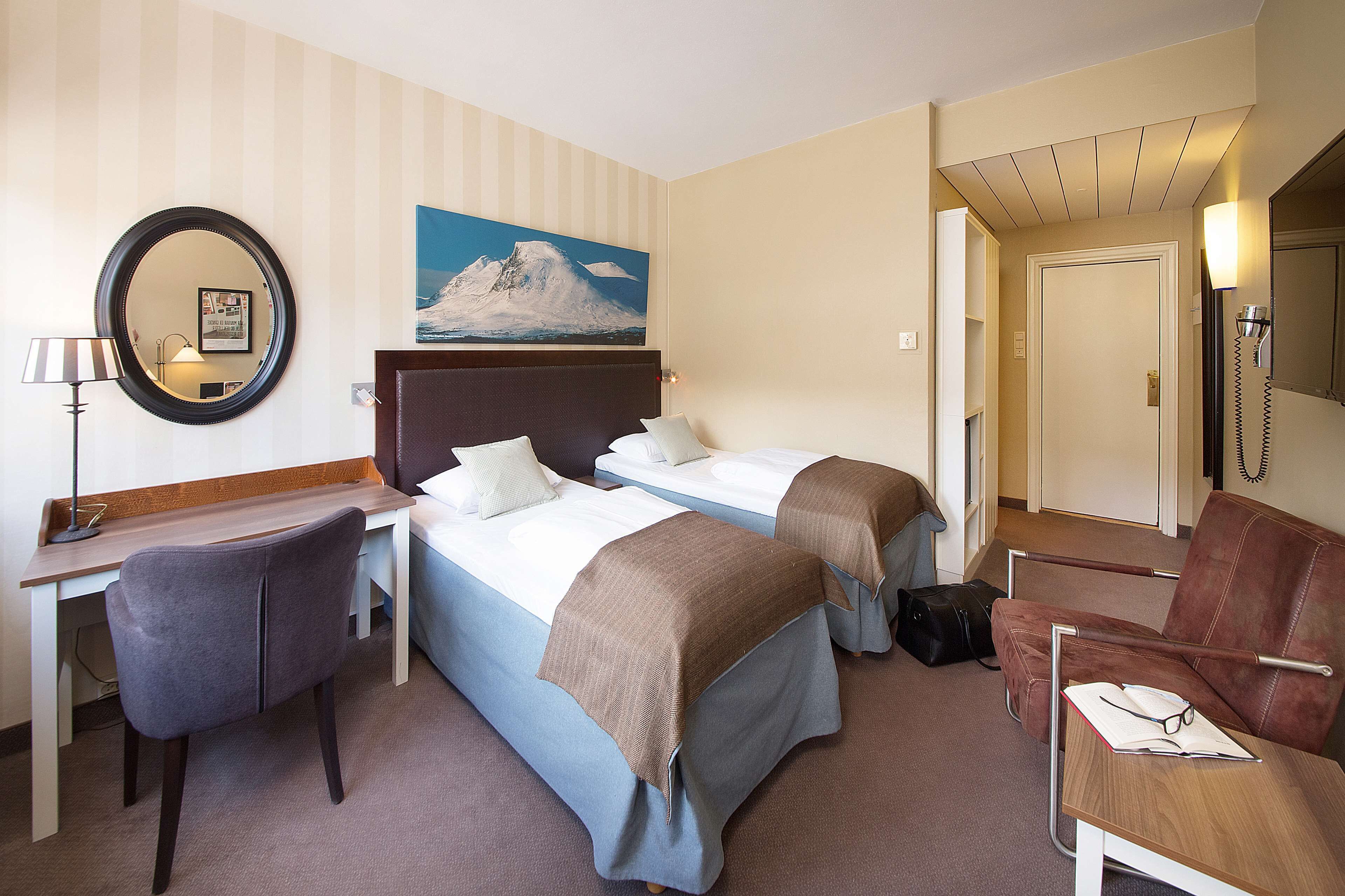 Quarto de hotel com duas camas, uma escrivaninha, uma cadeira, um espelho e um quadro de uma montanha.