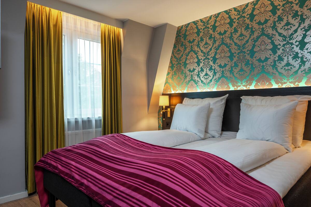 Quarto de hotel com uma cama, cortinas, parede estampada e mesa de cabeceira com abajur.