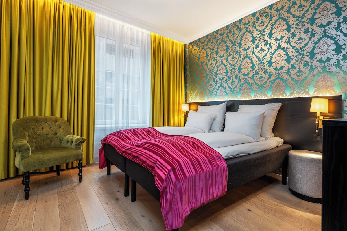 Quarto de hotel com cama, poltrona verde, mesa de cabeceira com abajur e papel de parede estampado.