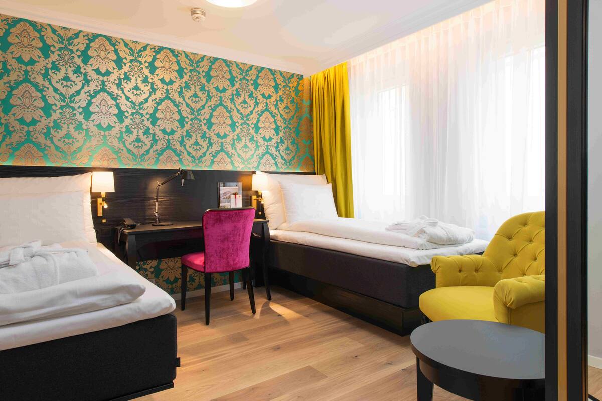 Quarto de hotel com duas camas, uma escrivaninha, uma cadeira e uma mesinha.