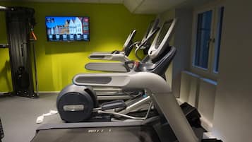 Uma academia com equipamentos de exercício, uma televisão exibindo um programa e uma parede verde.