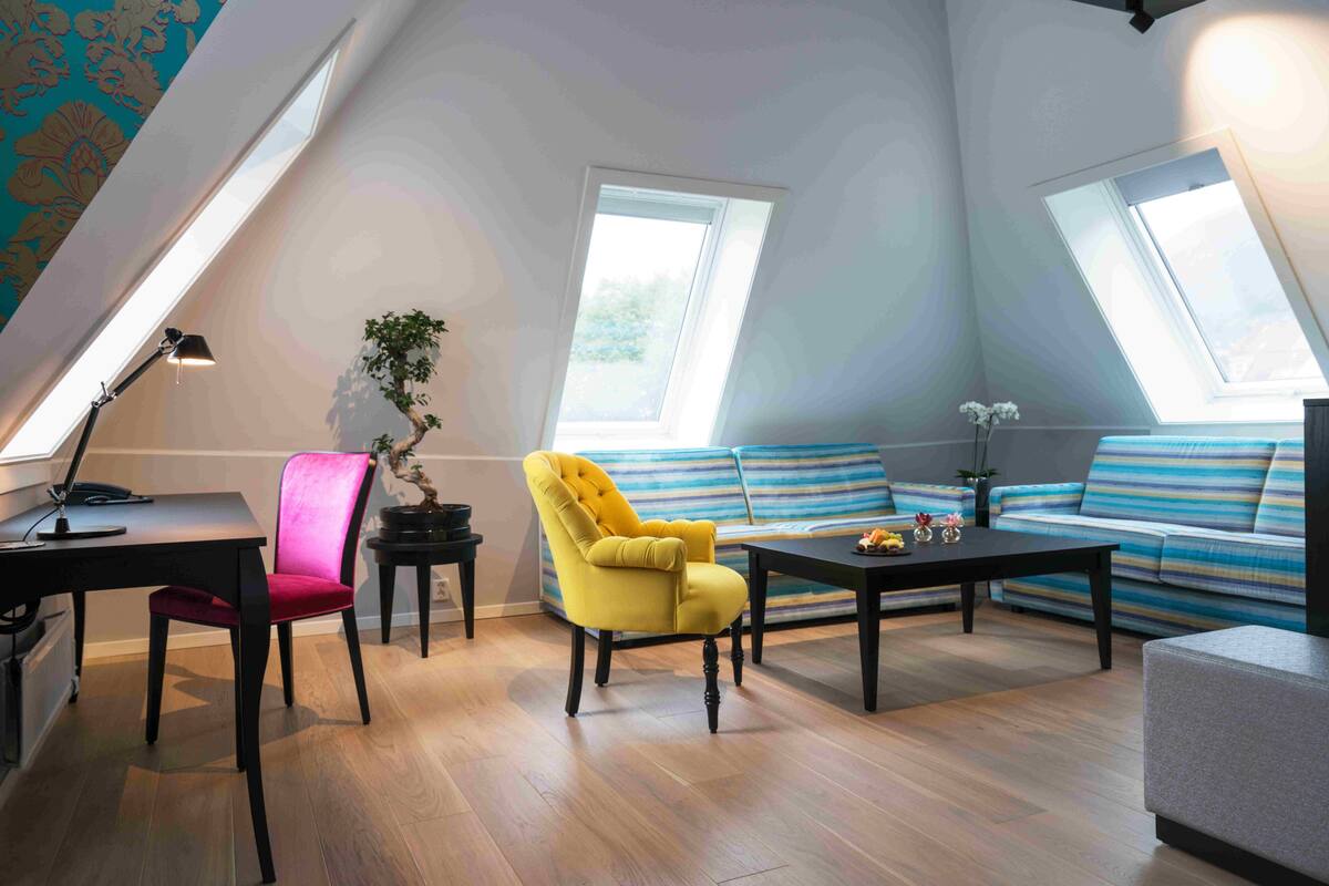 Um apartamento loft moderno com teto inclinado, claraboias, um conjunto de sofás, uma poltrona amarela, uma mesa de jantar e uma escrivaninha com uma luminária.