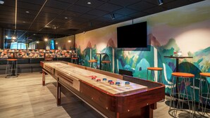 Uma área de bar com mesa de futebol de botão, banquetas e uma tela de televisão.