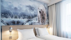 Quarto de hotel com um grande mural de urso polar na parede, cabeceira de madeira, duas almofadas brancas, mesa de cabeceira com abajur e janela com cortinas.
