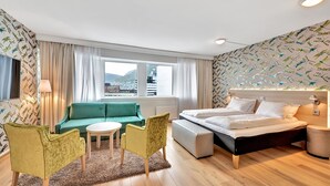 Quarto de hotel com cama, sofá verde, duas cadeiras amarelas, uma mesa pequena e vista para a paisagem urbana através de uma grande janela.