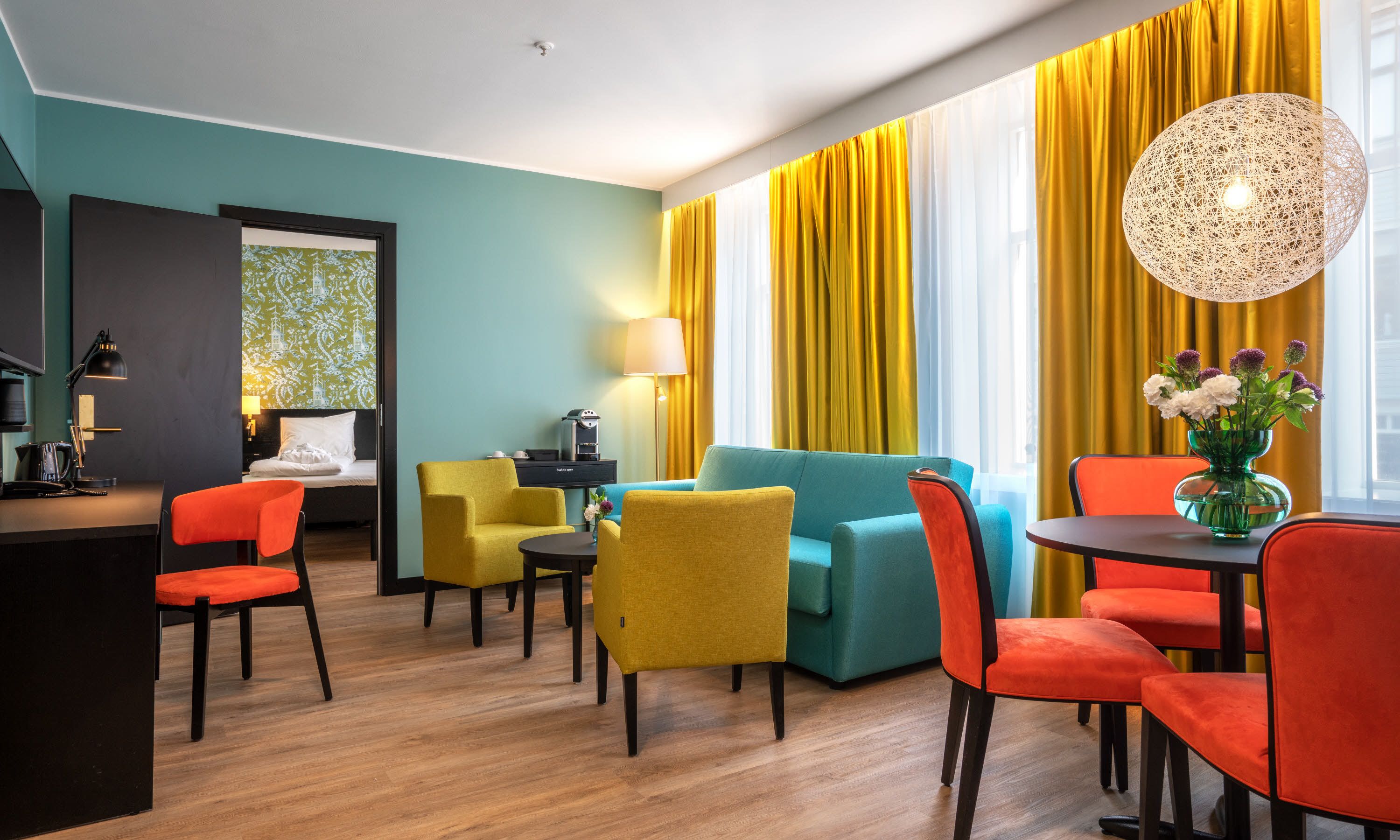 Quarto de hotel moderno com uma parede verde-água, cortina amarela, sofá azul e cadeiras laranja.