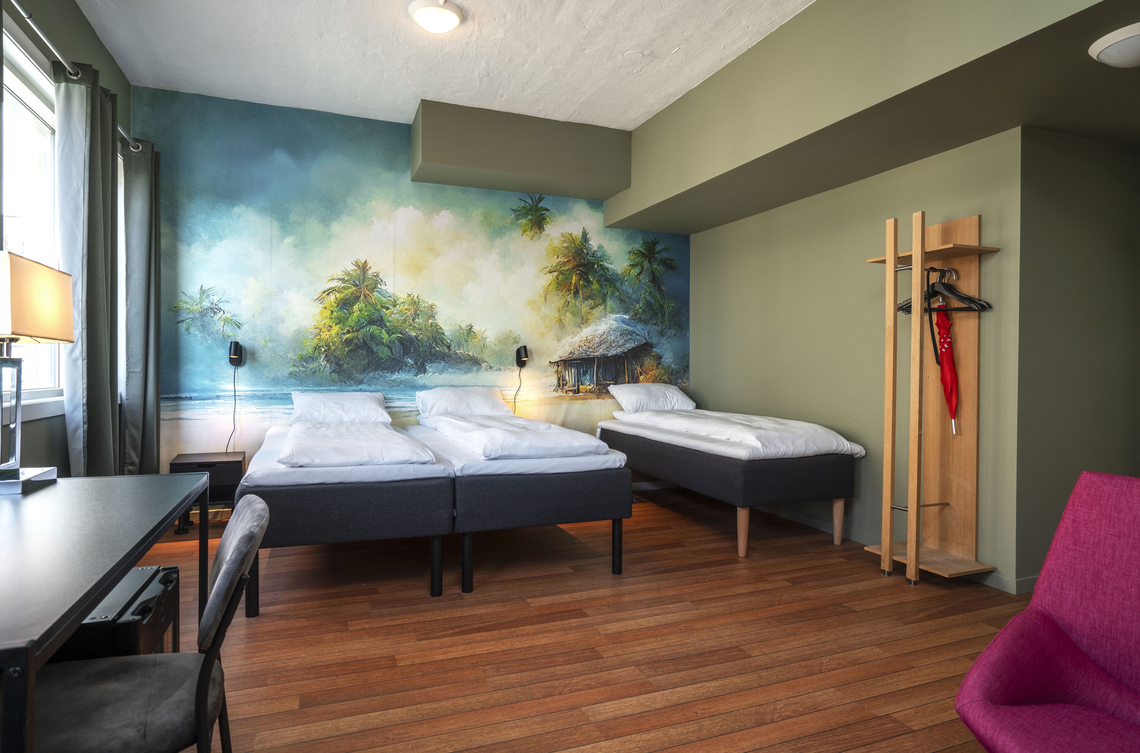 Quarto com duas camas, uma escrivaninha, uma cadeira, piso de madeira, painel com cena tropical e um cabideiro de madeira.