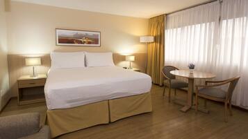 Quarto de hotel com uma cama grande, duas mesas de cabeceira com luminárias, uma mesa redonda pequena com uma cadeira e uma janela com cortinas.