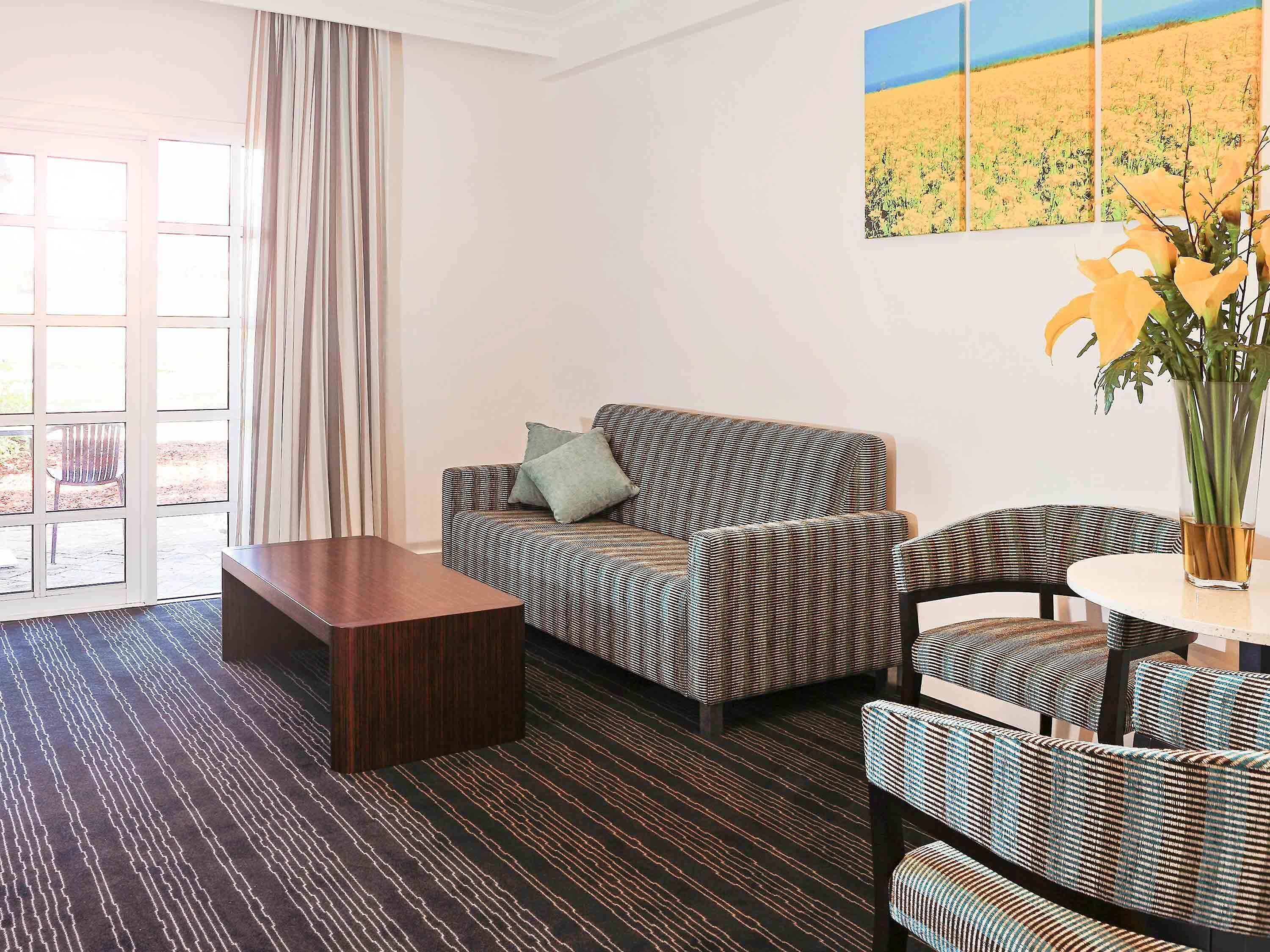 Sala de estar com sofá, mesa de centro e uma poltrona. Há uma grande janela com cortinas, um quadro na parede e um vaso com flores sobre uma mesa.