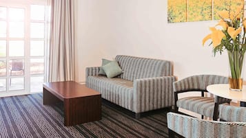 Sala de estar com sofá, mesa de centro e uma poltrona. Há uma grande janela com cortinas, um quadro na parede e um vaso com flores sobre uma mesa.