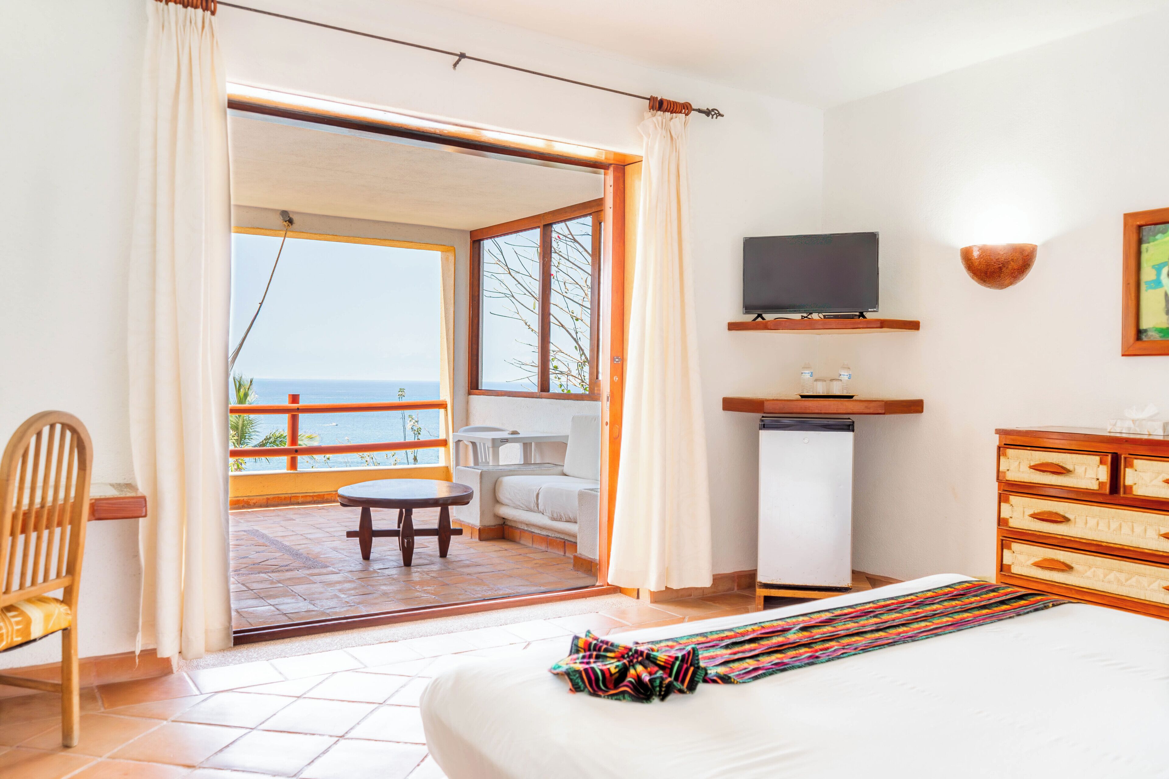 Quarto de hotel com cama, televisão, penteadeira, escrivaninha, cadeira e varanda com vista para o mar.