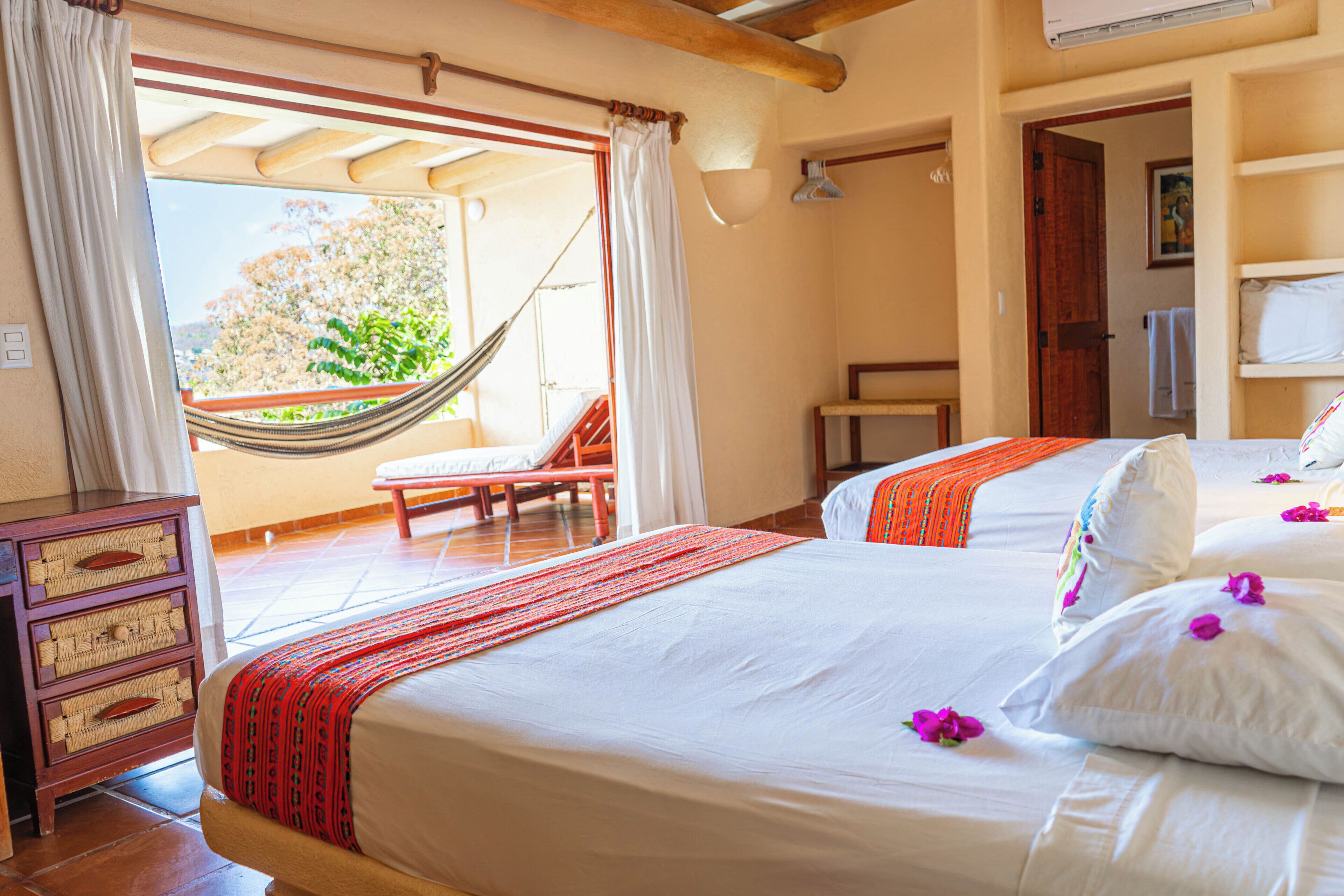 Quarto de hotel com duas camas, uma rede, uma cadeira de madeira e vista para o verde lá fora.