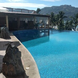Uma piscina azul cristalina sem fim com vista para uma montanha e palmeiras ao fundo.