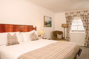 Quarto de hotel bem arrumado, com uma cama grande, uma poltrona, uma luminária e uma janela com cortinas.