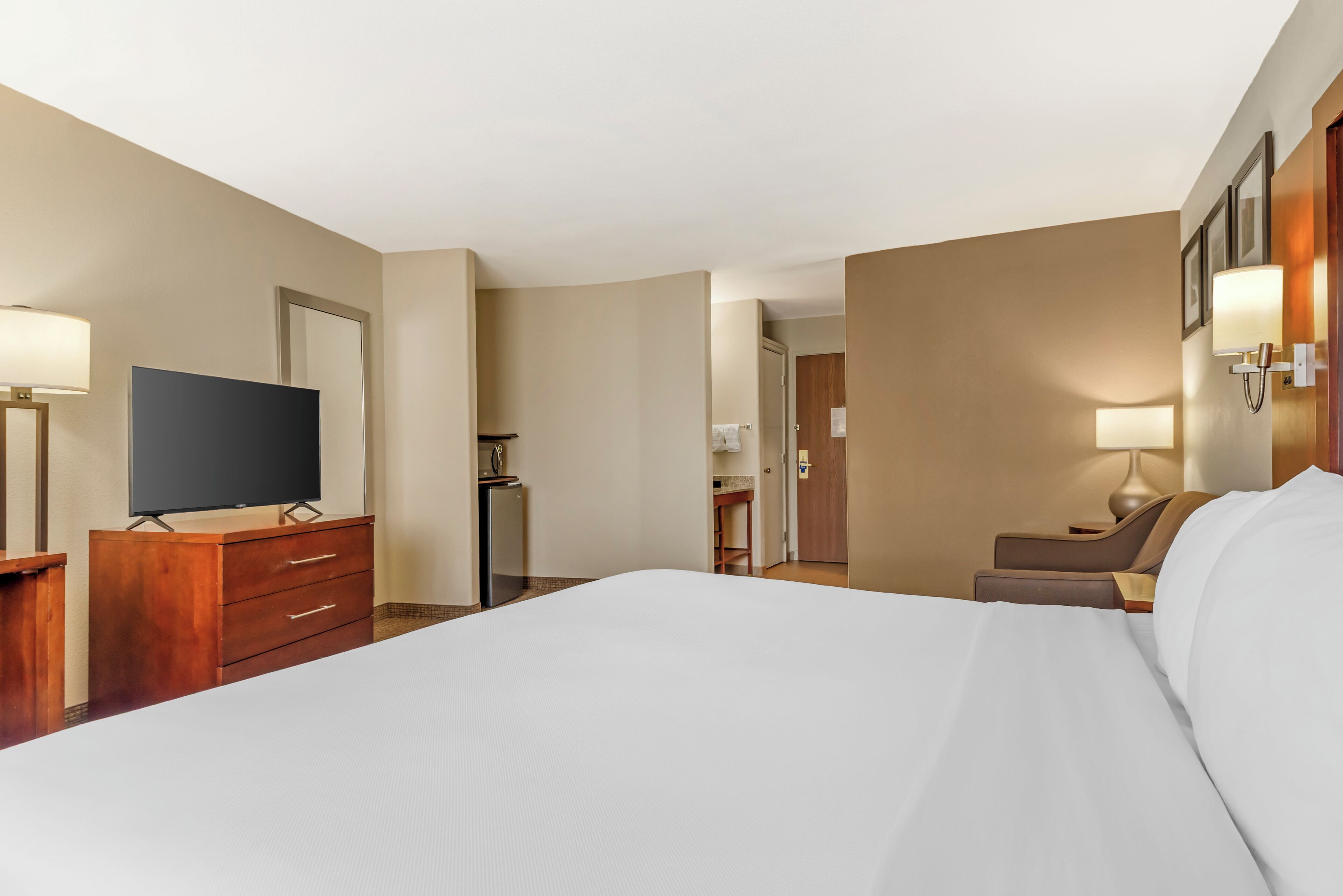 Quarto de hotel com uma cama grande, uma TV sobre um aparador de madeira, uma mesa de trabalho com cadeira e uma porta que dá para outro quarto.