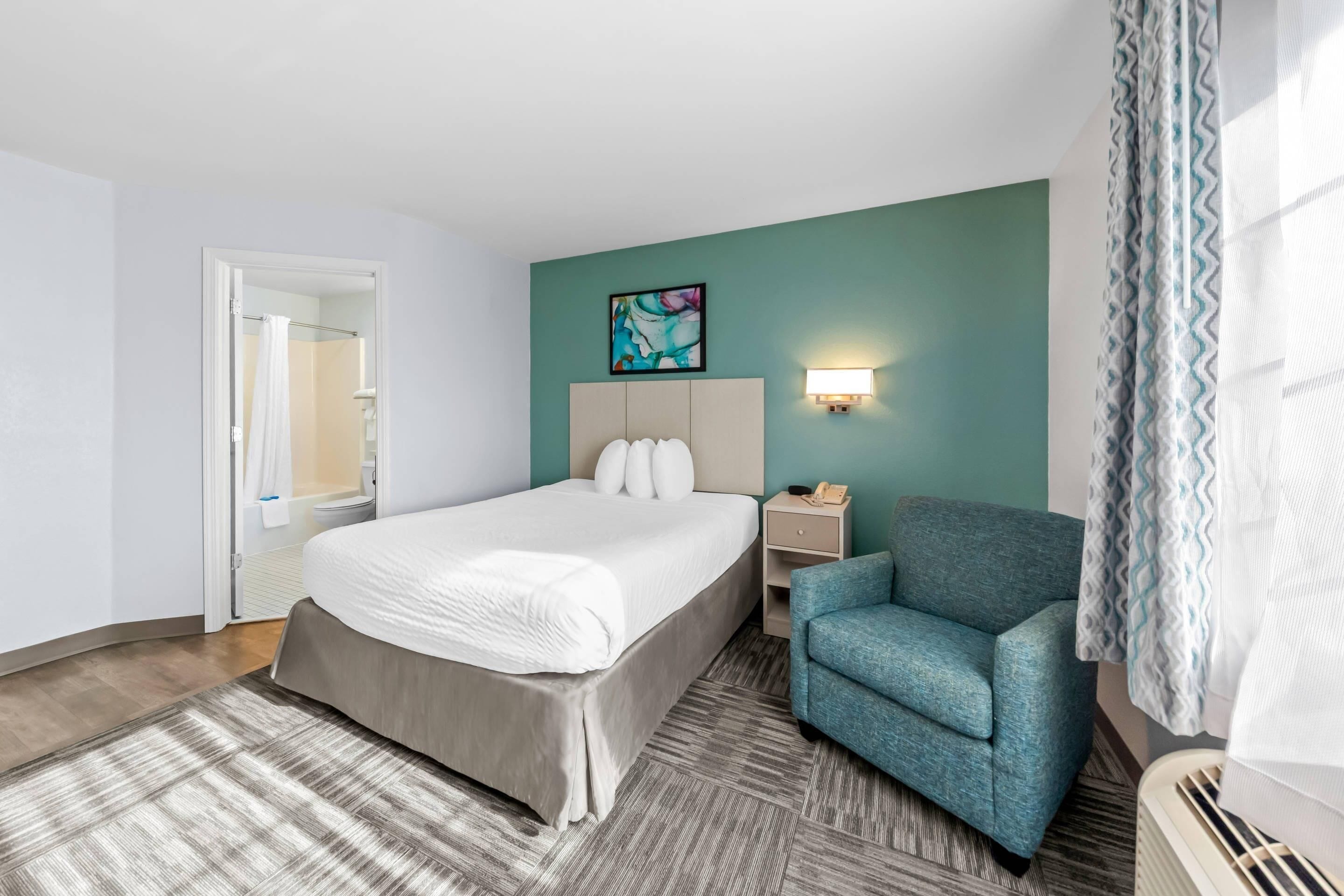 Quarto de hotel com cama, cadeira, mesa de cabeceira, quadro e janela com cortinas.