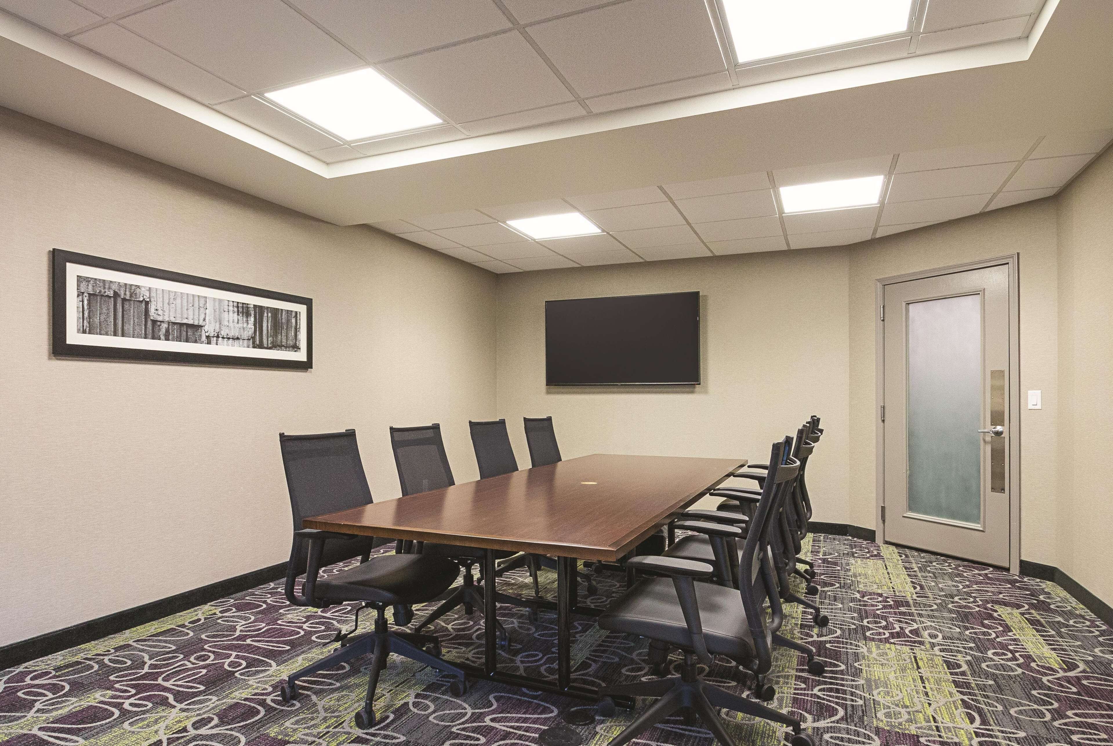 Uma sala de conferências com uma mesa comprida, várias cadeiras, uma tela de projetor e uma porta.