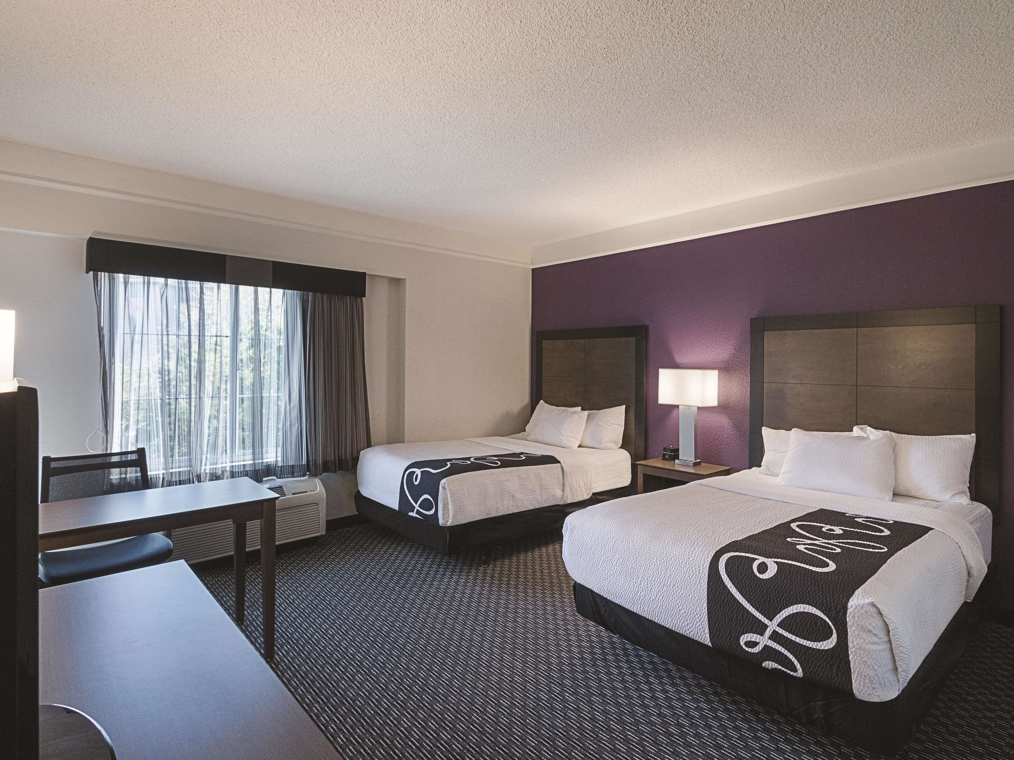 Quarto de hotel com duas camas, uma escrivaninha, uma cadeira, um abajur e uma janela com cortinas.