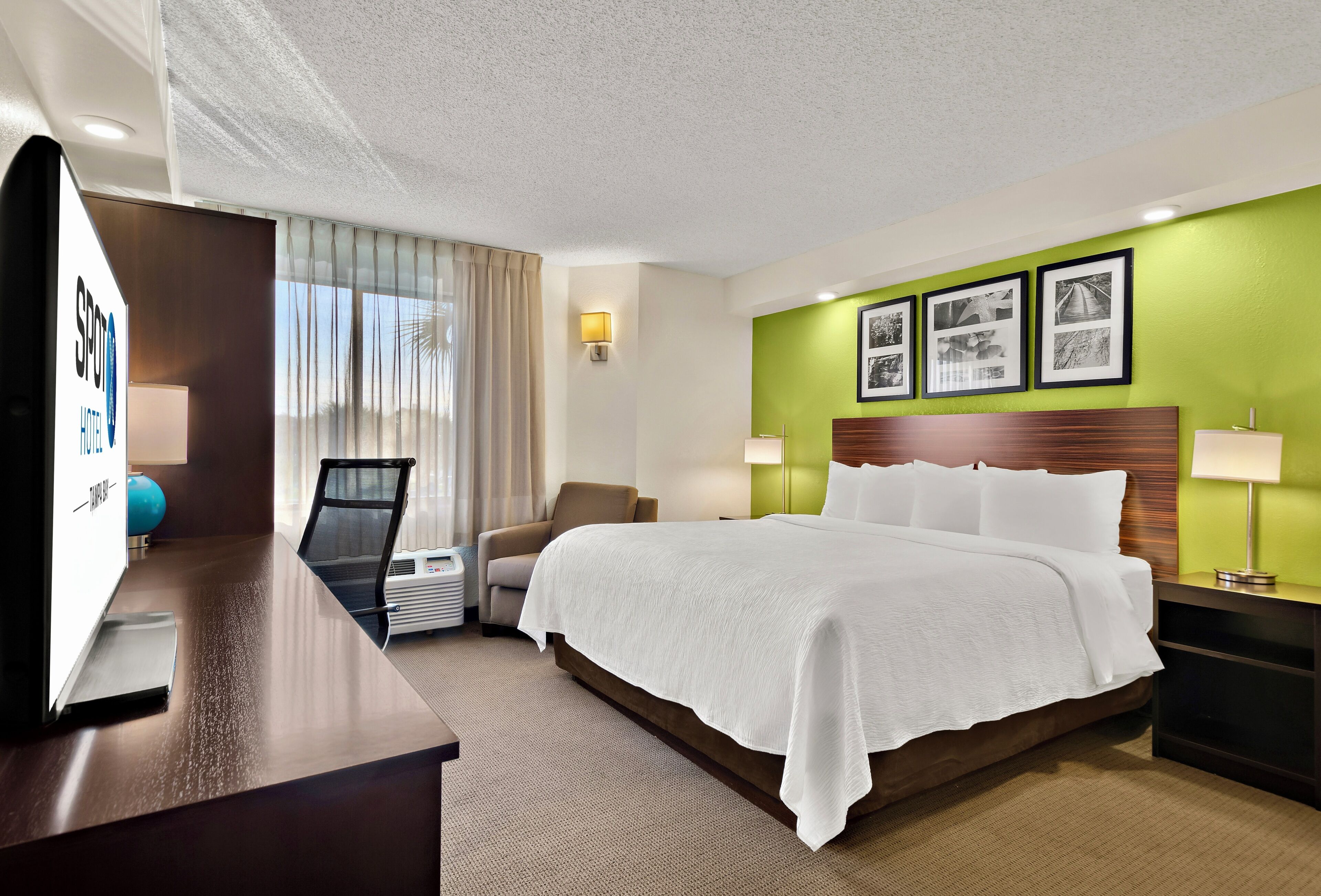 Quarto de hotel com cama, escrivaninha, cadeira, janela com cortinas, luminária na parede e quadros emoldurados.