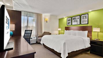 Quarto de hotel com cama, escrivaninha, cadeira, janela com cortinas, luminária na parede e quadros emoldurados.