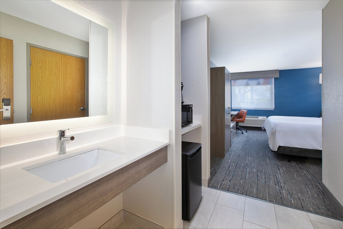 Quarto de hotel moderno com uma pia de banheiro grande, um refrigerador compacto, cama com cobertura branca e parede azul.