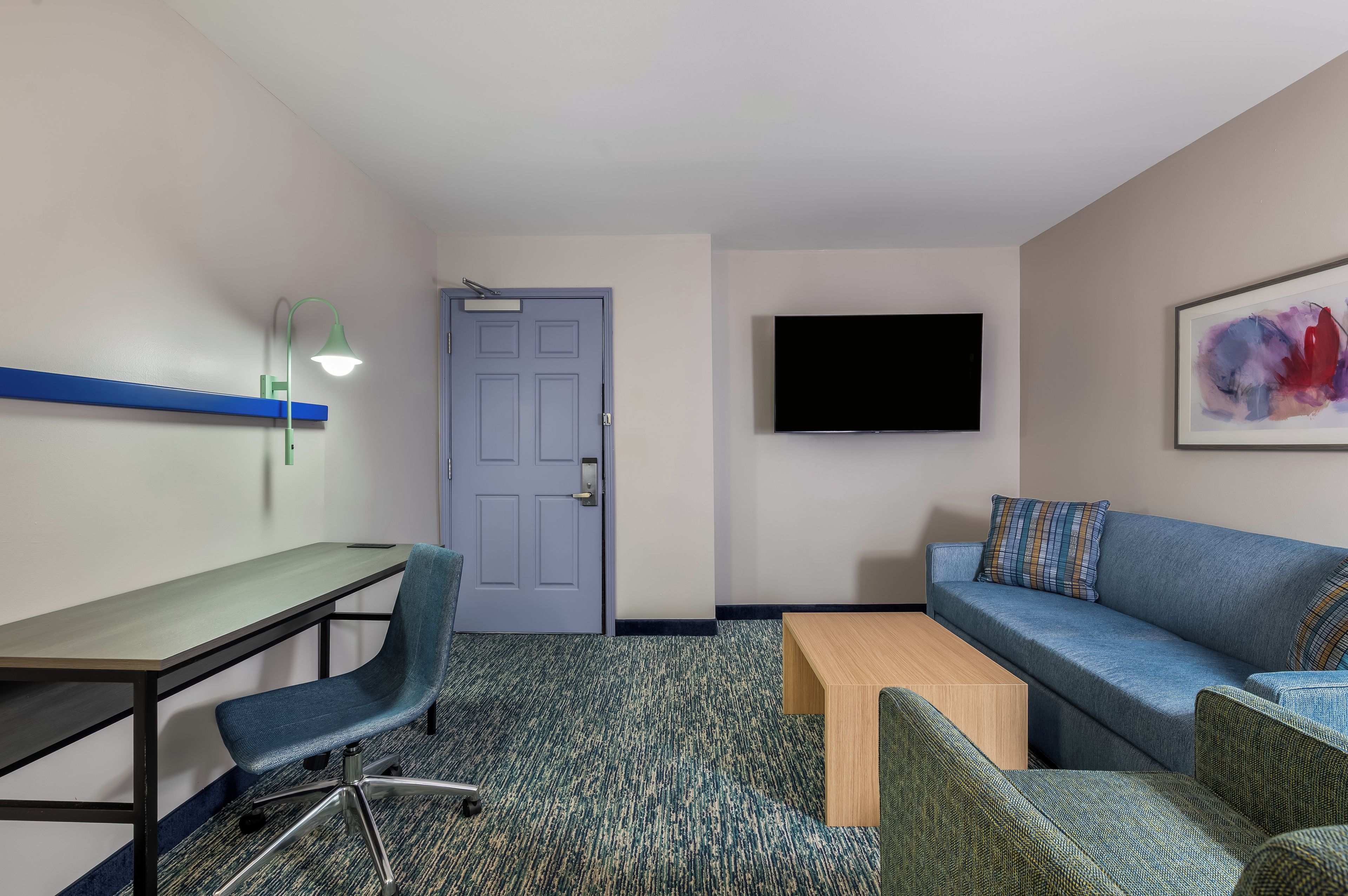 Quarto de hotel com escrivaninha, cadeira, sofá e televisão de tela plana.