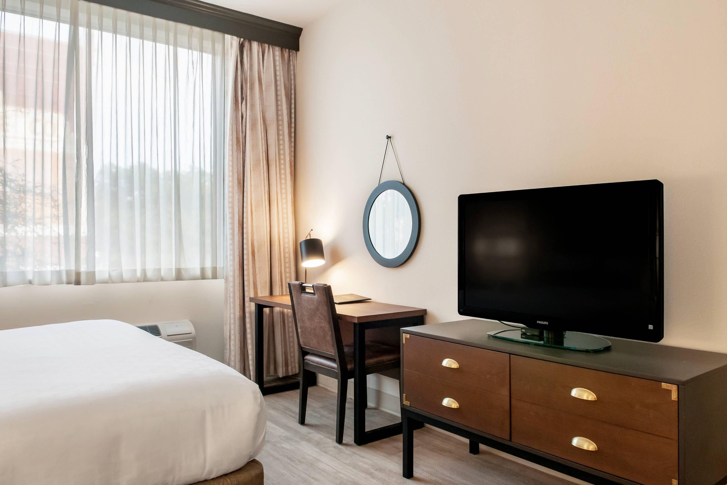 Quarto de hotel com uma cama, uma escrivaninha com cadeira, televisão num móvel e uma janela com cortinas.