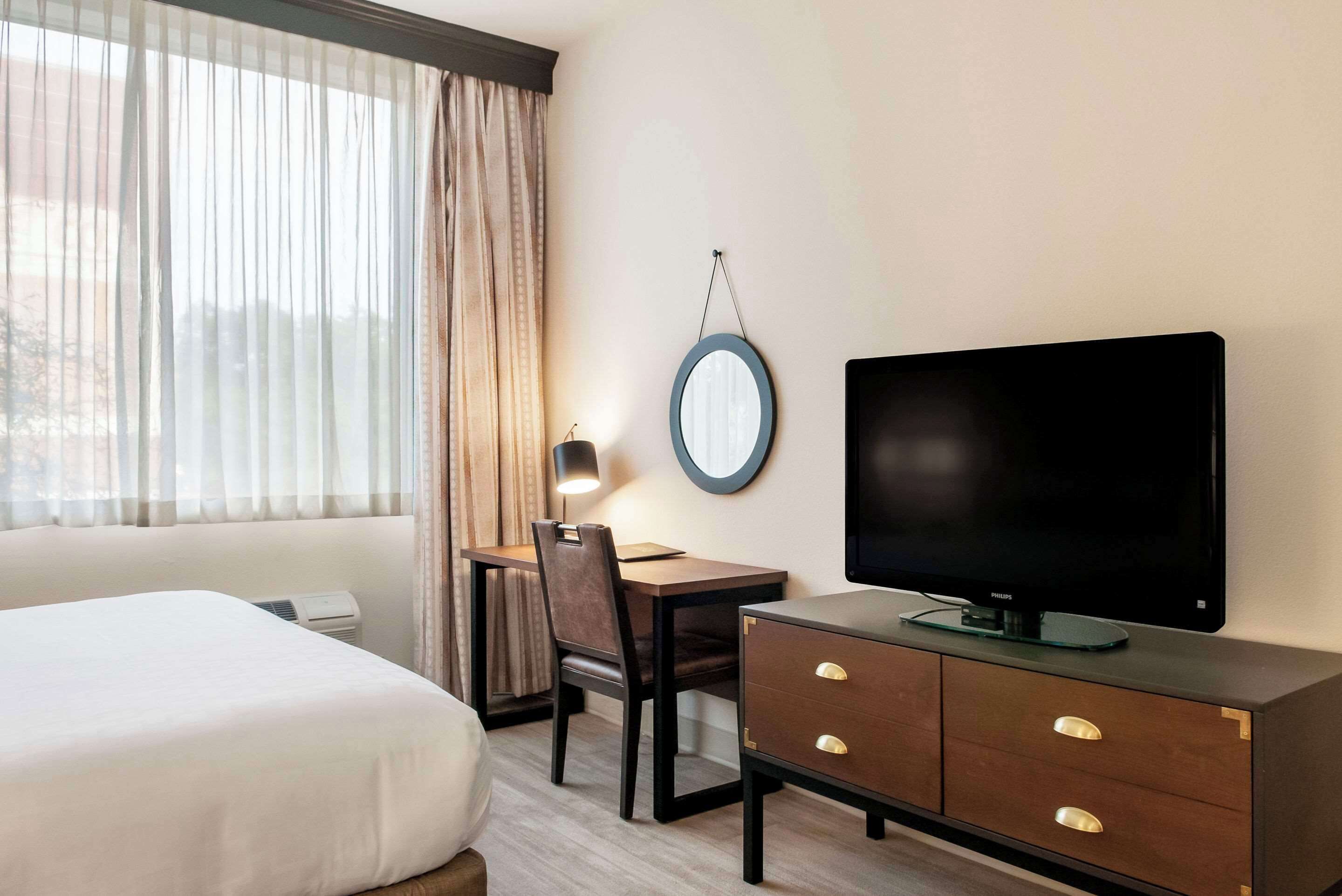 Quarto de hotel com cama, escrivaninha com cadeira, televisão e espelho na parede.