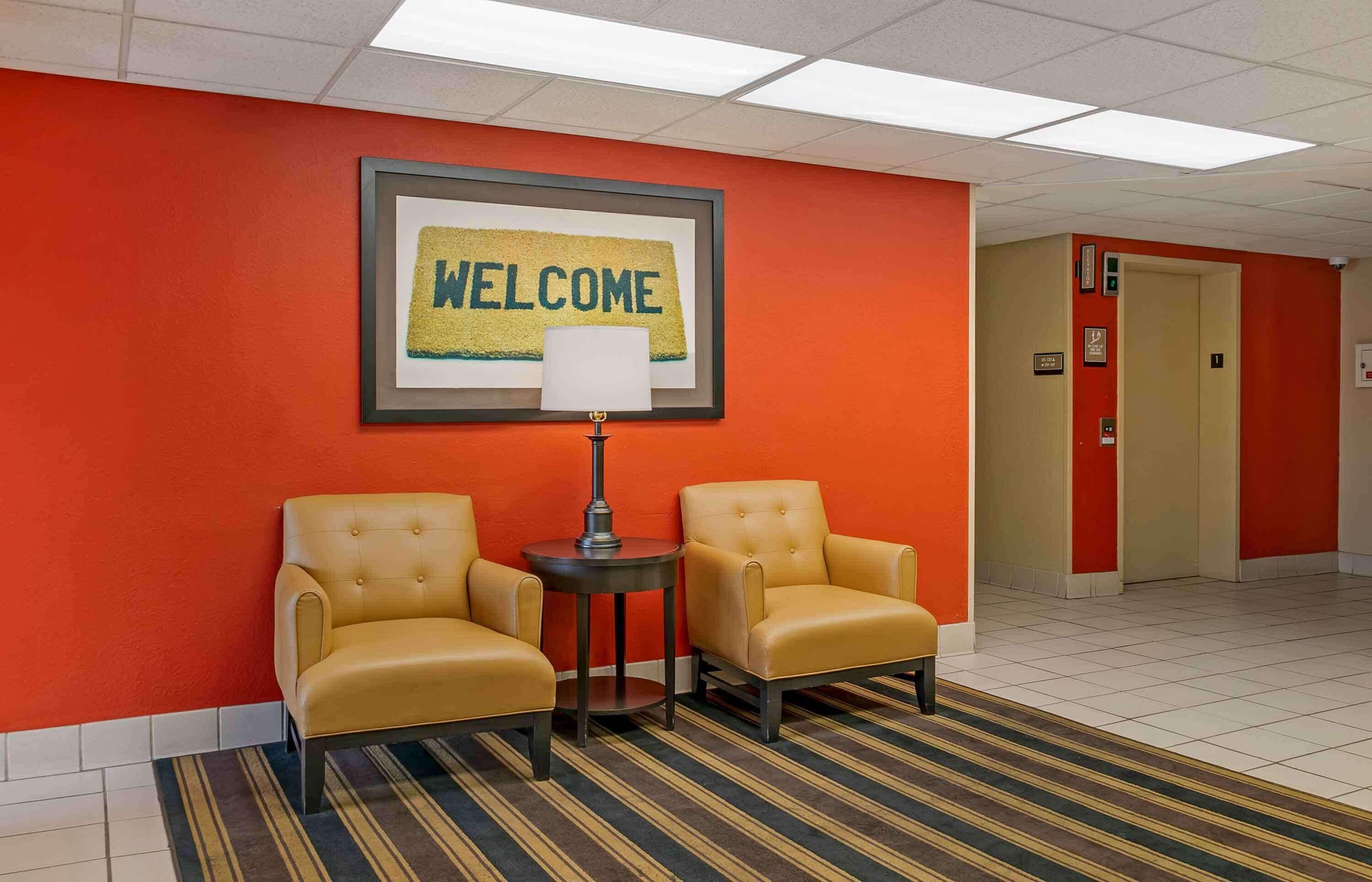 Um saguão de hotel com um letreiro 'WELCOME', duas poltronas de couro, uma mesinha com abajur e um tapete listrado.