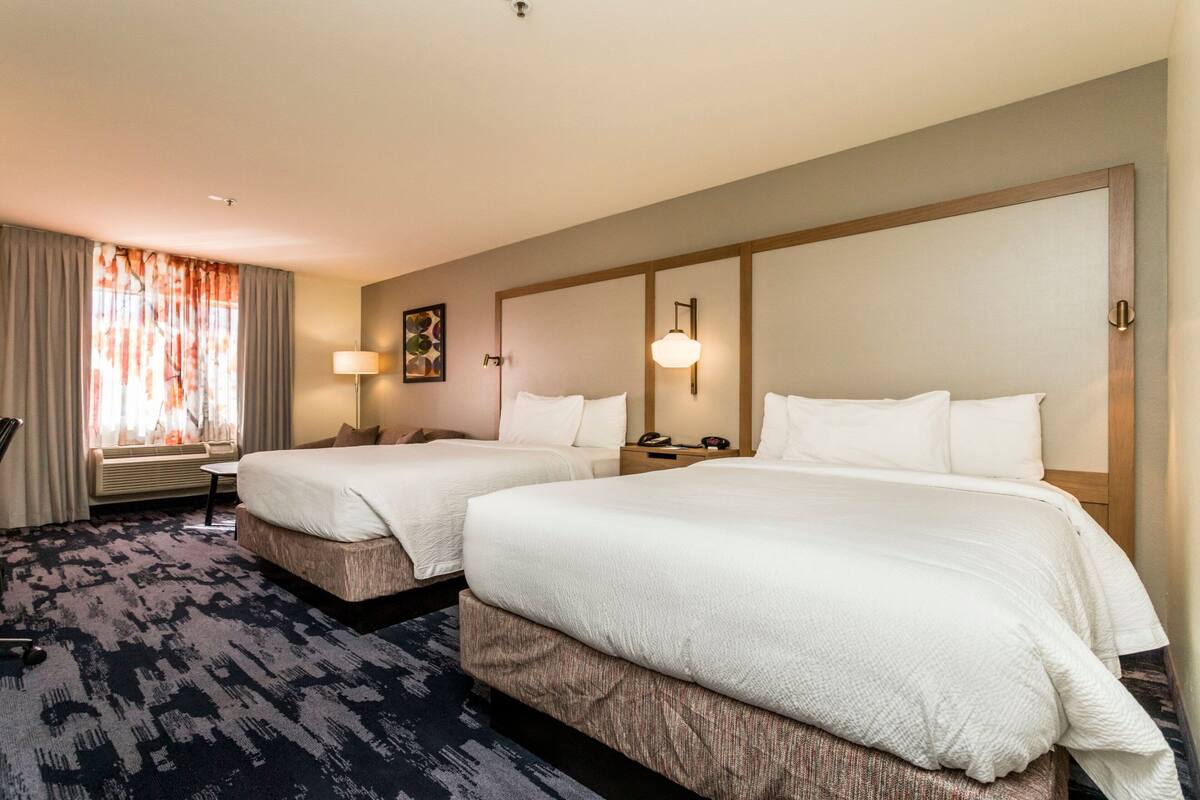 Quarto de hotel com duas camas, uma janela grande com cortinas, uma luminária fixada na parede e uma pequena mesa.