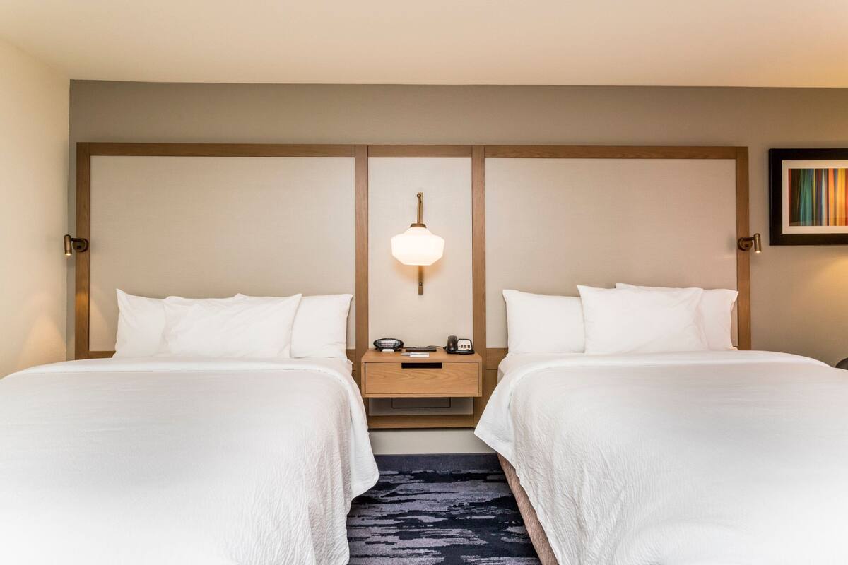 Quarto de hotel com duas camas, uma mesa de cabeceira com abajur e um quadro na parede.