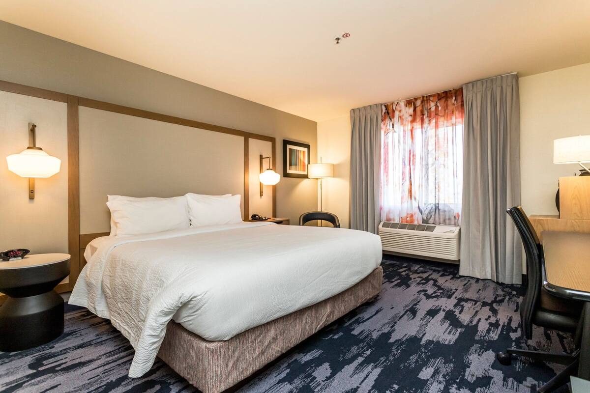 Quarto de hotel com uma cama grande, duas mesas de cabeceira, uma escrivaninha com cadeira, uma janela com cortinas e um aparelho de ar condicionado fixado na parede.
