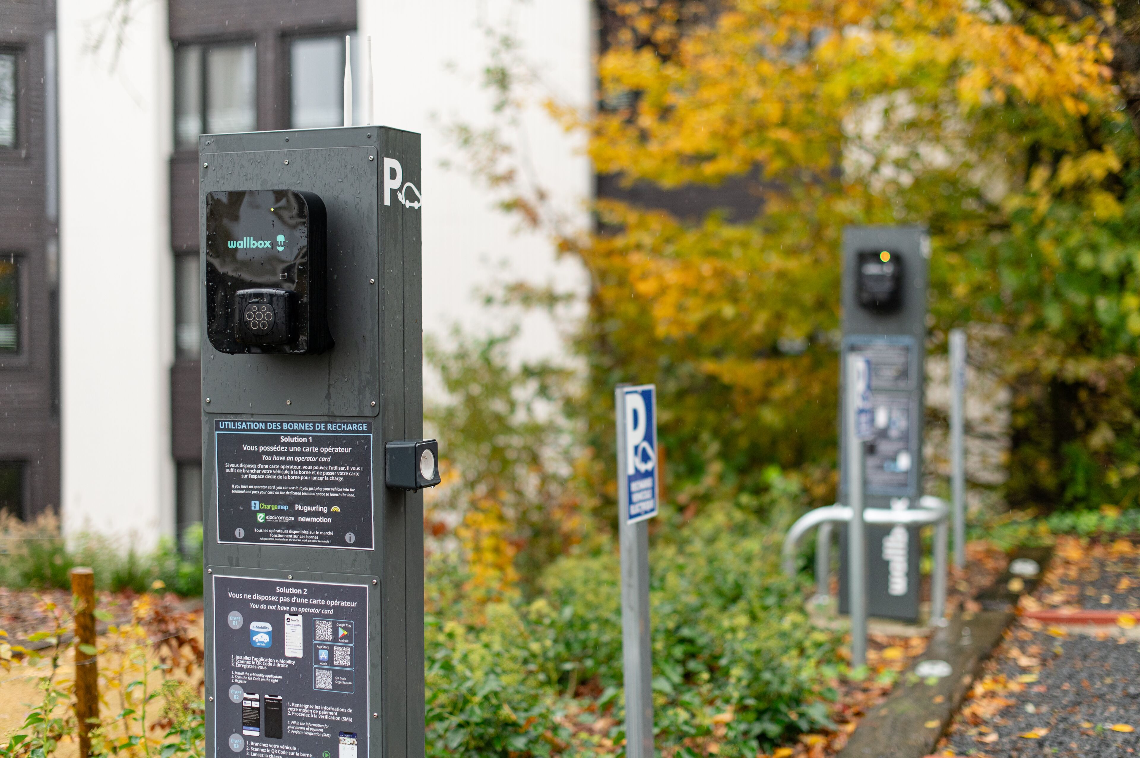 Um parquímetro com instruções em francês, uma placa de estacionamento e um edifício ao fundo.