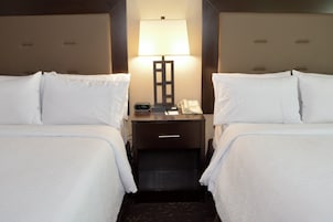 Quarto de hotel com duas camas, uma mesa de cabeceira com abajur, telefone e relógio digital.