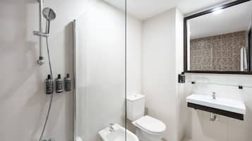 Uma casa de banho moderna com cabine de duche em vidro, sanita branca, bidé e lavatório com prateleira de vidro acima.