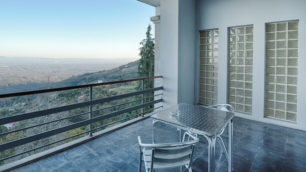 Uma varanda com mesa e cadeiras, vista para o campo e um edifício com janelas grandes.