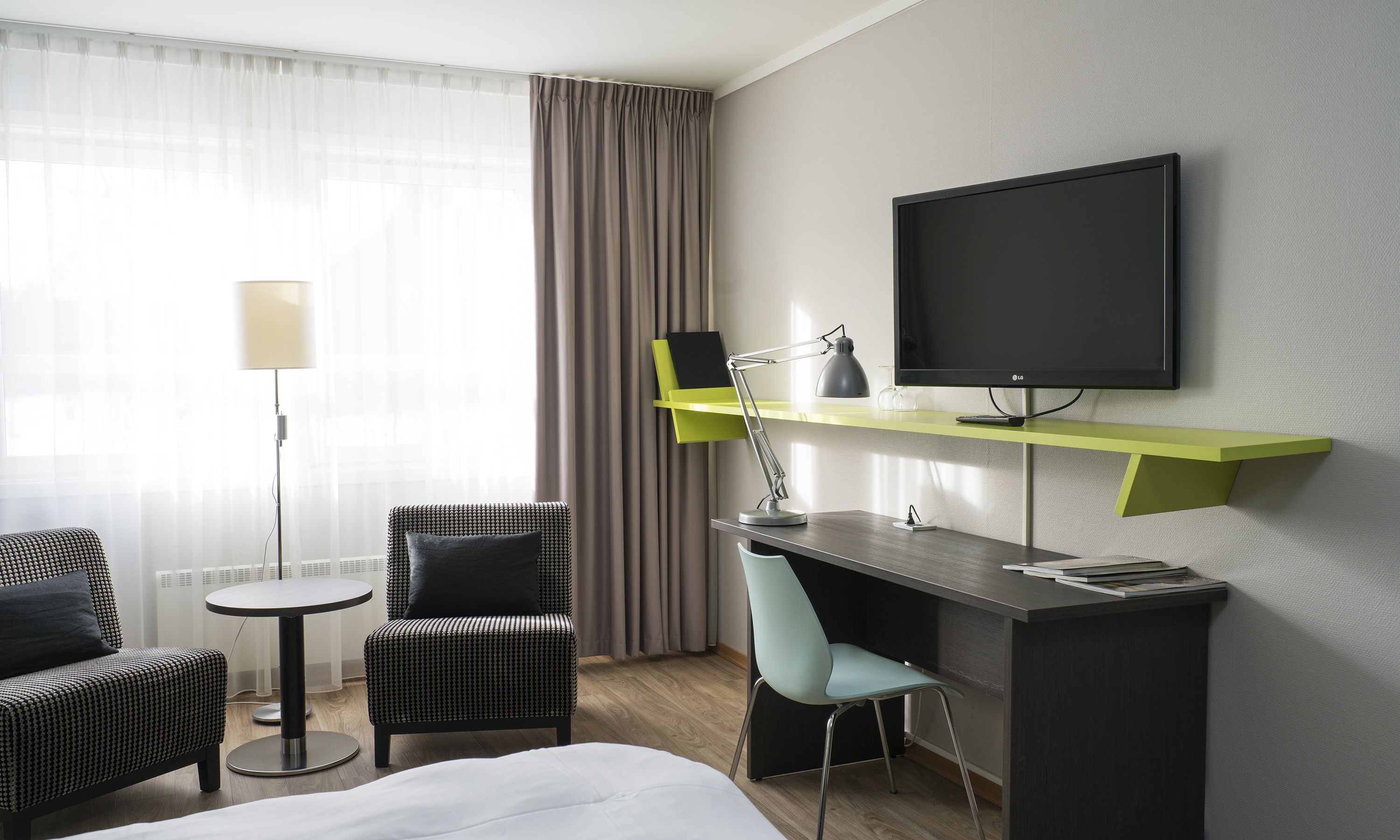 Quarto de hotel com cama, escrivaninha, cadeira, televisão e abajur.