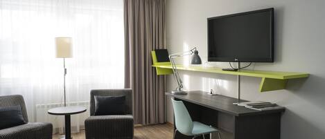 Quarto de hotel com cama, escrivaninha, cadeira, televisão e abajur.
