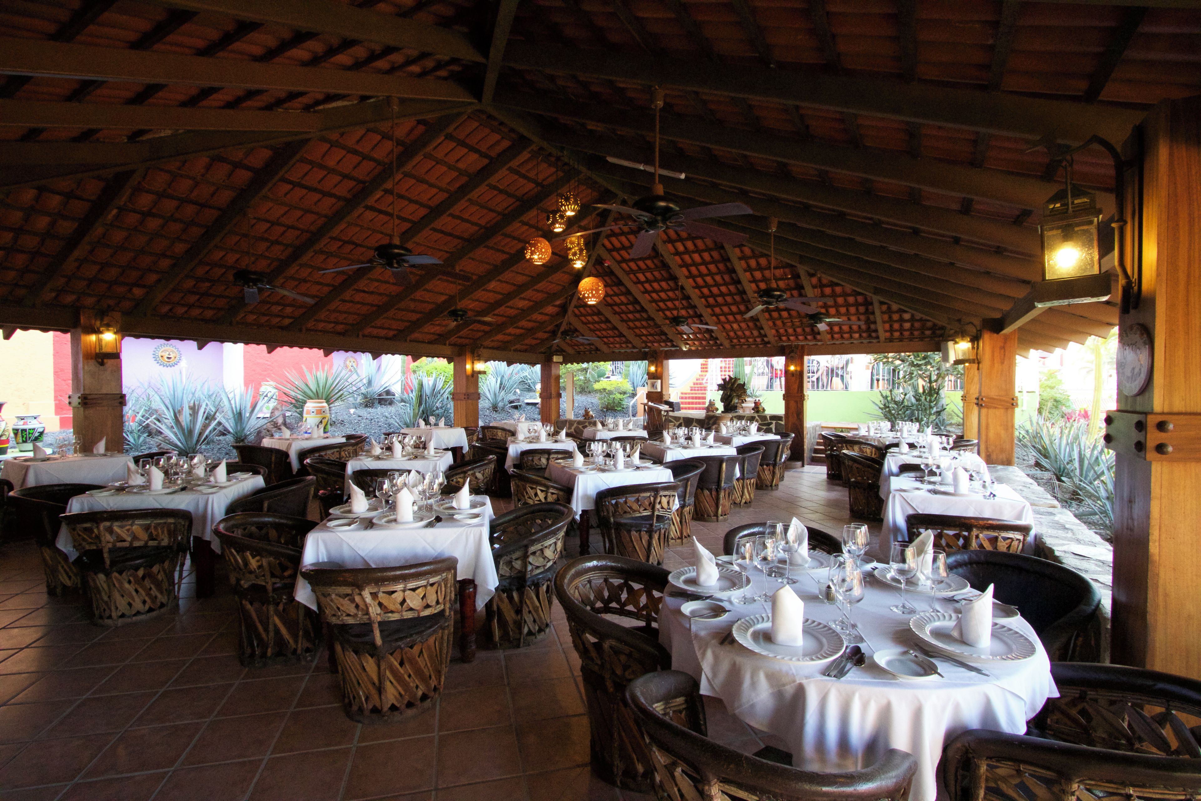 Restaurante ao ar livre com várias mesas preparadas para refeições, rodeado por cadeiras de vime e um chão de azulejos.