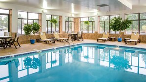 Piscina interna com espreguiçadeiras e mesas, amplas janelas e plantas em vasos.