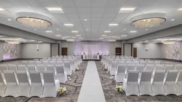 Uma sala de conferências ampla e bem iluminada, com fileiras de cadeiras brancas, um corredor central e lustres.