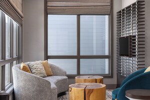 Uma sala de estar moderna com um sofá cinza curvo, uma poltrona azul e duas mesas laterais de madeira. Uma janela grande com persianas e uma TV montada em um painel metálico são visíveis.