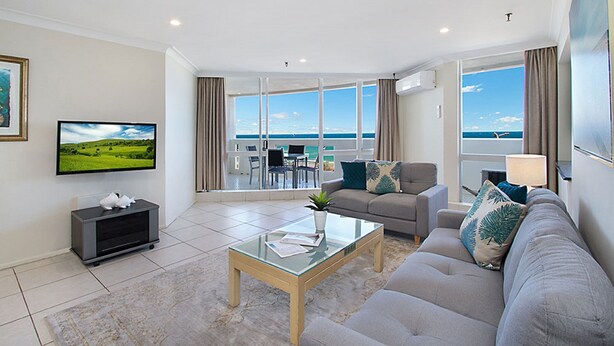 Sala de estar moderna com sofá, mesa de centro de vidro, TV de tela plana e vista para o mar através de amplas janelas.