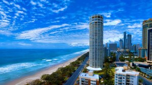 Paisagem costeira urbana com um edifício cilíndrico imponente próximo à praia, cercado por arranha-céus e um céu azul límpido.