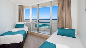 Quarto de hotel com duas camas, uma janela ampla com vista para o mar e uma varanda com cortinas.